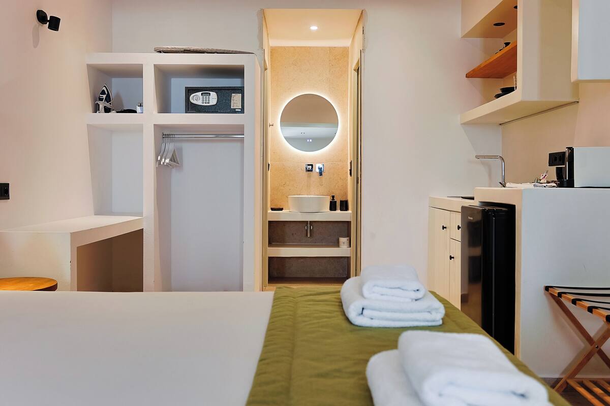Quarto de hotel com cama, colcha verde, cama bem arrumada com toalhas brancas, uma pequena mesa, espelho fixado na parede, pia, geladeira e prateleira com artigos de higiene pessoal.