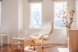 Quarto moderno com duas cadeiras brancas, piso de madeira e janela com vista para árvores.