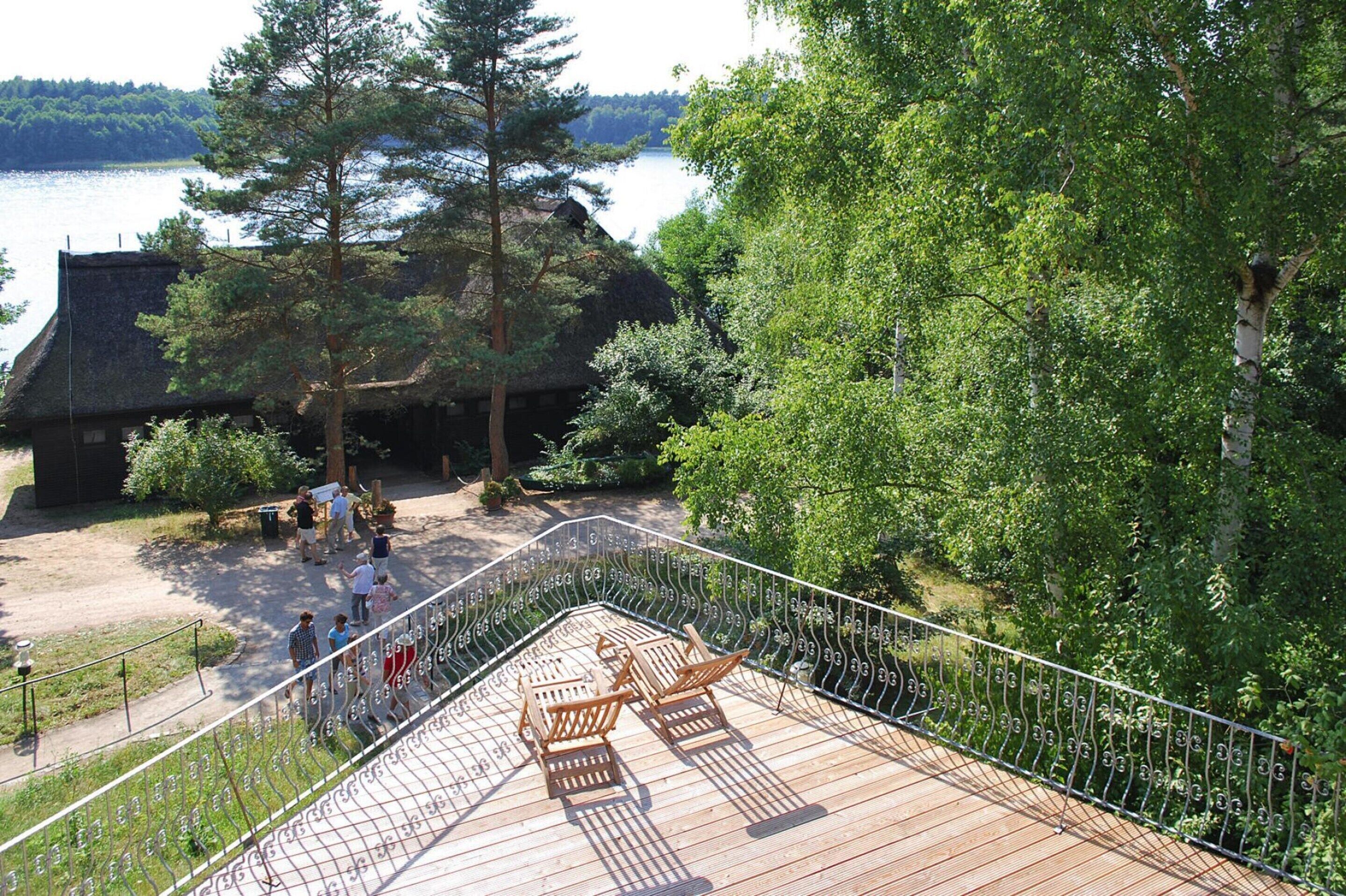 Um deck de madeira com cadeiras oferece vista para um lago, cercado por árvores e um prédio com telhado de palha.