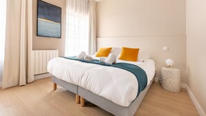 Cama bem arrumada com roupa de cama branca e azul-turquesa, dois travesseiros amarelos e um cobertor azul-turquesa. Um radiador branco é visível na parede, e uma pequena luminária branca está sobre uma mesa redonda ao lado da cama.