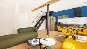 Uma sala moderna com um sofá, uma mesa de centro redonda e uma cadeira amarela. Uma escada leva a uma cozinha com uma parede azul e uma área de jantar com cadeiras amarelas.