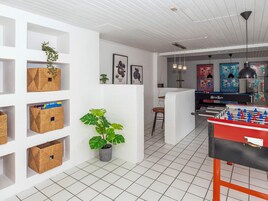 Quarto moderno com paredes brancas, mesa de futebol e televisão. Há prateleiras com cestos e um vaso de planta.