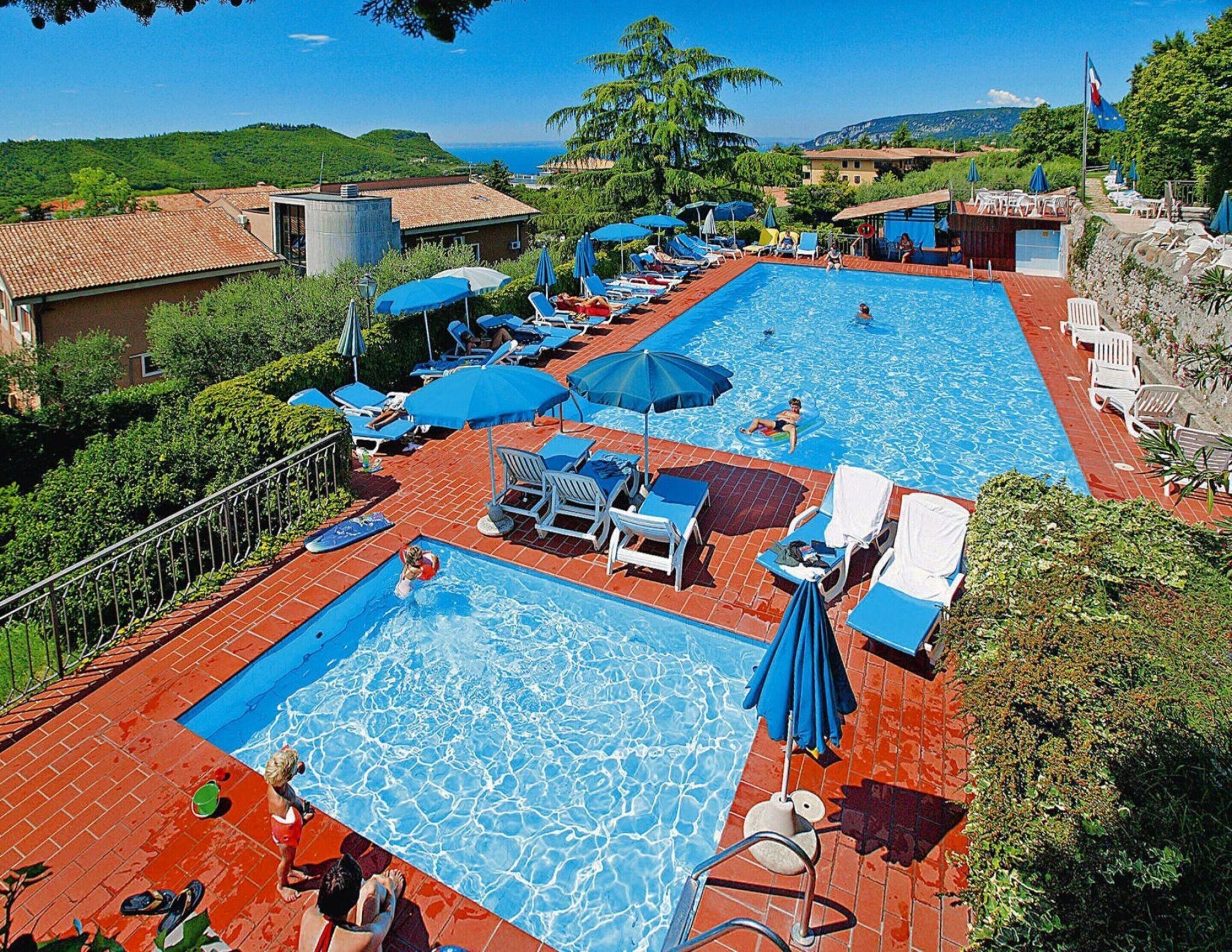 Uma grande piscina ao ar livre com várias espreguiçadeiras e guarda-sóis, rodeada por vegetação e edifícios.