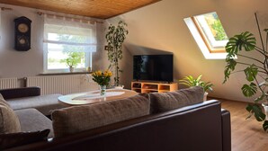 Uma sala de estar acolhedora com teto de madeira, televisão de tela plana, sofá, mesa de centro com flores e uma grande janela com vista para o verde.