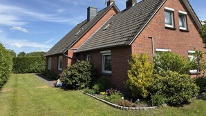 Uma casa de dois andares de tijolo, com telhado inclinado, chaminé visível e um jardim bem cuidado com diversas plantas e gramado.