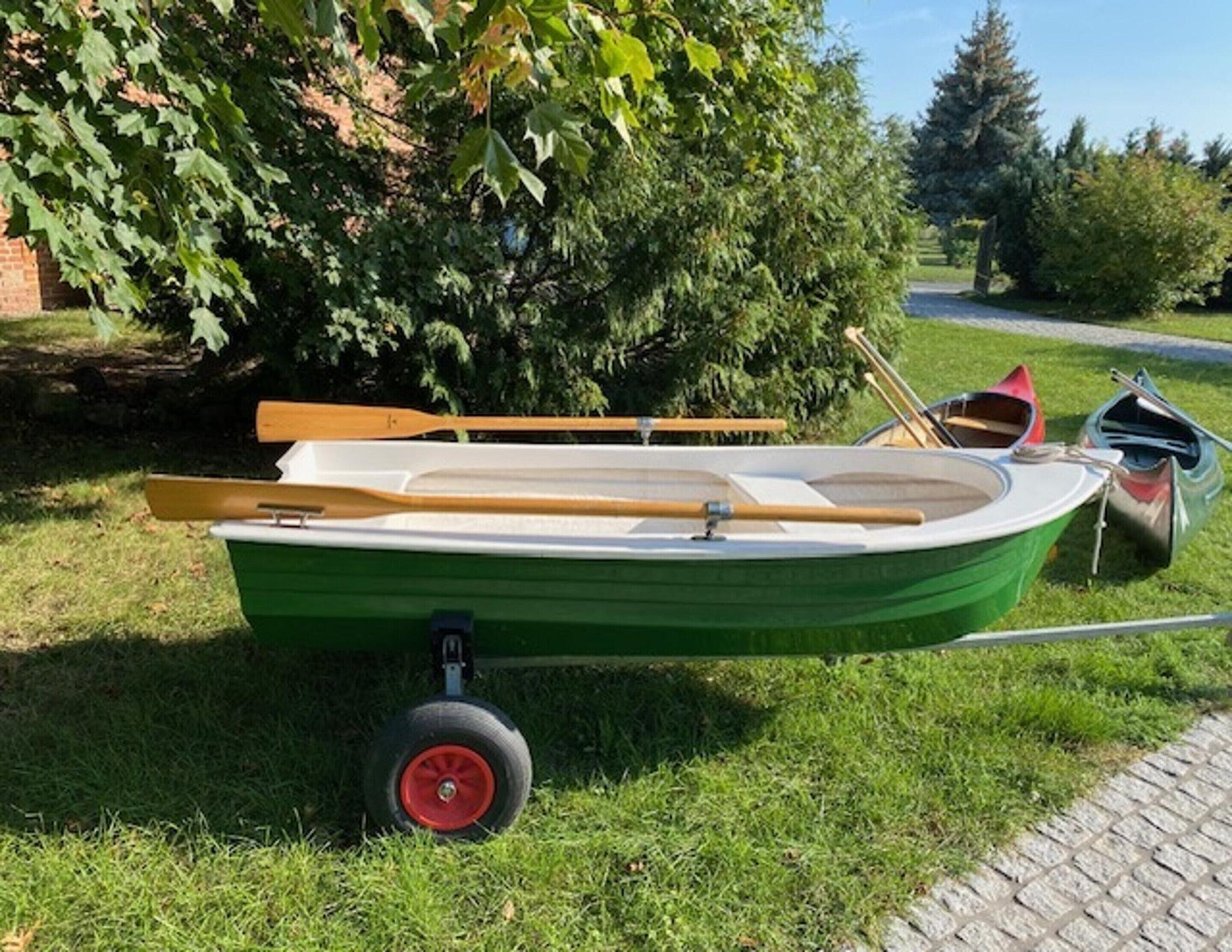 Um pequeno barco verde com um banco de madeira e remo, preso a um reboque com uma roda vermelha, estacionado na relva ao lado de um sebe.