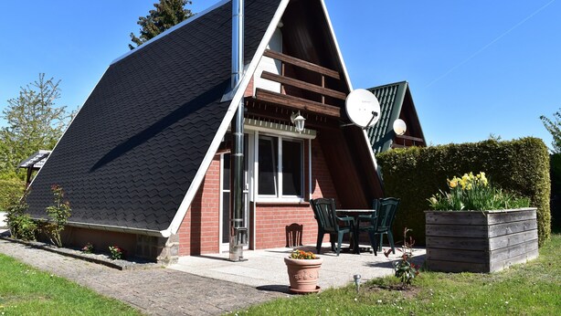 Uma casa moderna com telhado inclinado, antena parabólica e jardim com flores e um vaso de madeira.
