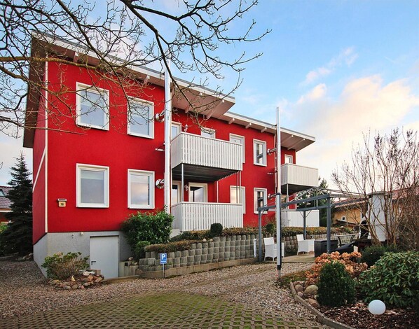 Um edifício residencial vermelho com dois andares, varanda, entrada pavimentada e pequeno jardim.