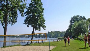 Uma quadra de voleibol de praia com rede, rodeada por árvores e um lago ao fundo.