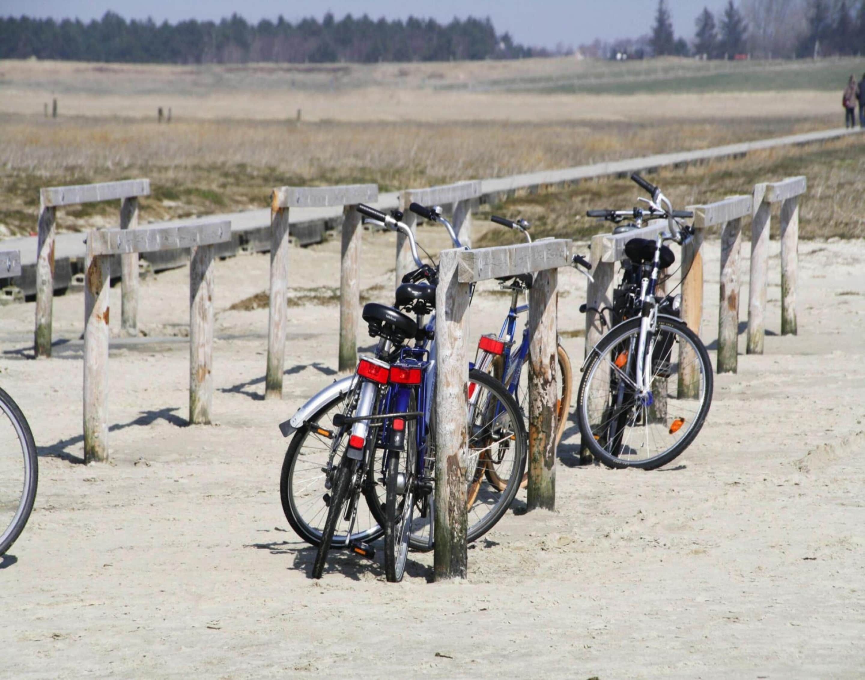 Várias bicicletas estacionadas em uma praia arenosa, com uma cerca de madeira ao fundo.