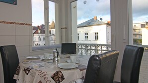 Uma área de refeições com uma mesa posta para dois, uma cafeteira e vista para prédios através de amplas janelas.