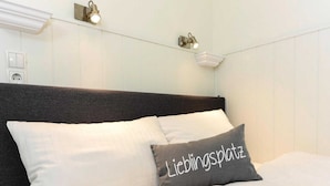 Cama com almofadas brancas e cinzas, cabeceira escura e luminárias fixadas na parede.