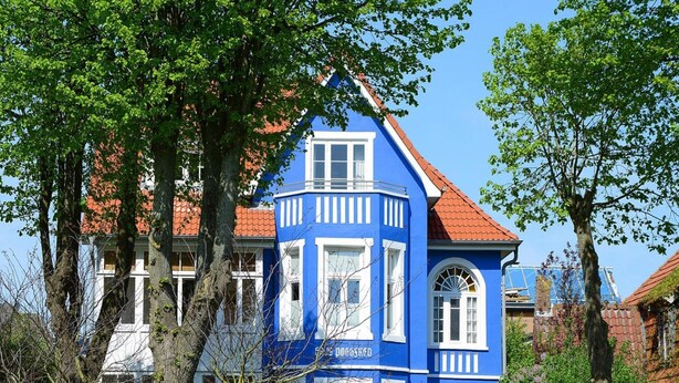 Um edifício azul com várias janelas e telhado vermelho, rodeado por árvores e vegetação.
