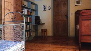 Quarto com uma cama de ferro, guarda-roupa de madeira, estante de livros e um banquinho pequeno.