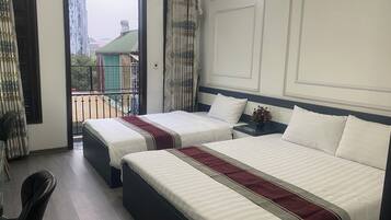 Quarto de hotel com duas camas, uma pequena mesa, uma cadeira e vista para um prédio através de uma porta aberta.