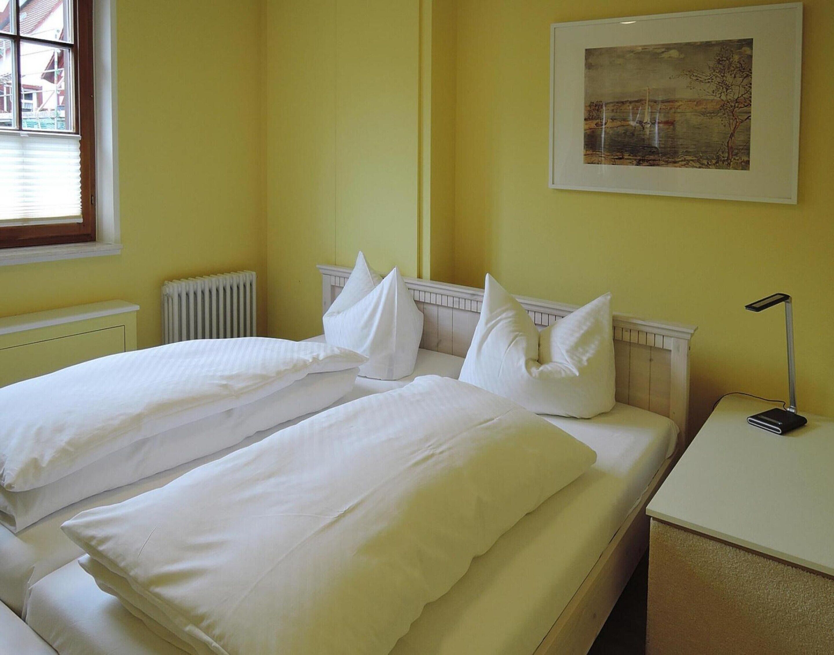 Uma cama de casal com lençóis e almofadas brancas, uma mesa de cabeceira com abajur e um quadro na parede.