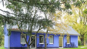 Uma casa térrea azul com varanda frontal, um pequeno jardim e uma árvore em frente.