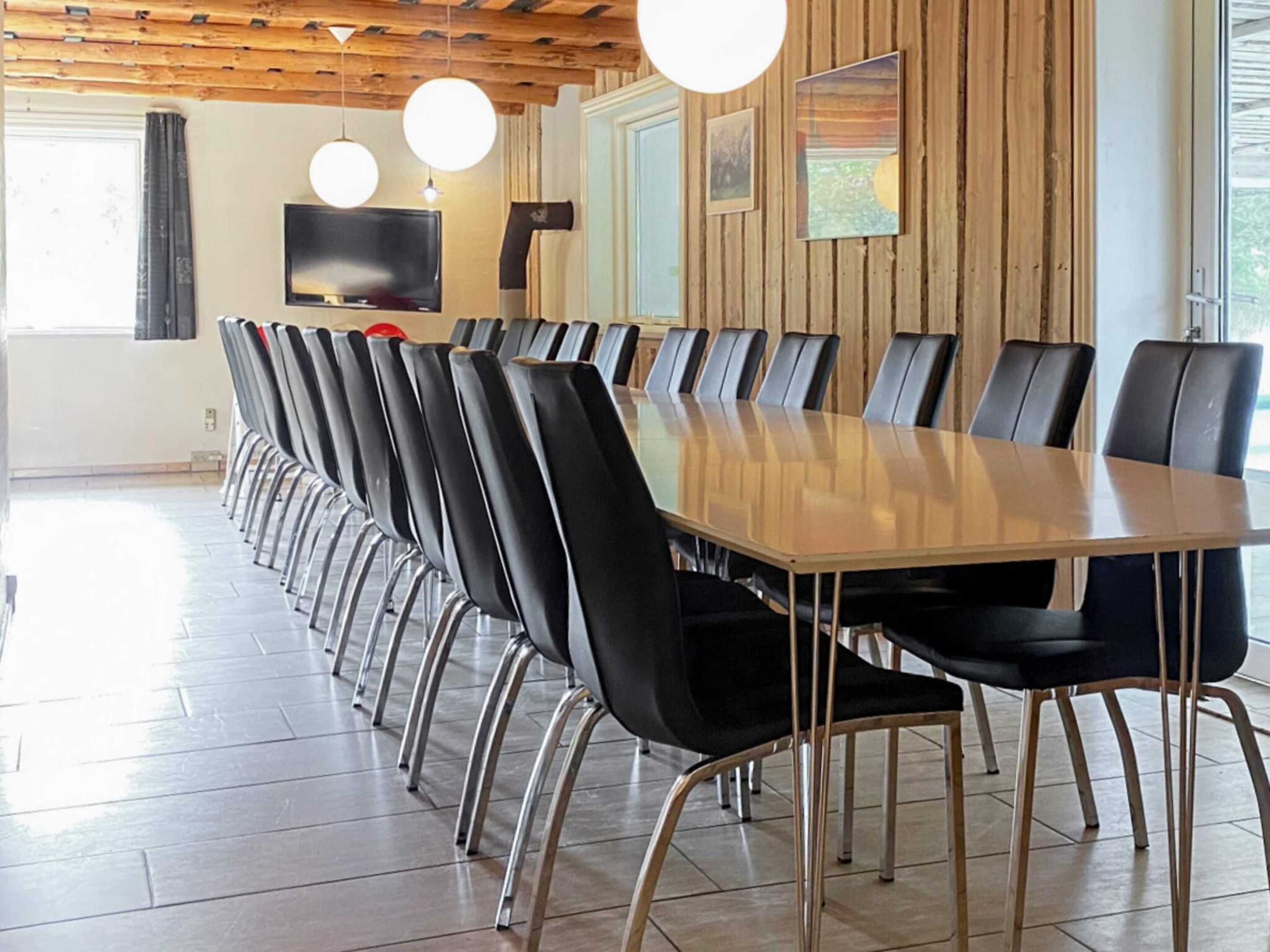 Uma sala de conferências com uma mesa comprida e cadeiras, paredes com painéis de madeira, uma televisão e iluminação pendente.
