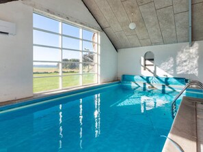 Piscina interna com janelas de vidro transparente, ventilador de teto e vista para uma paisagem verde.