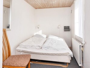 Uma cama de solteiro com edredom branco, uma cadeira de madeira, uma mesinha e um radiador.