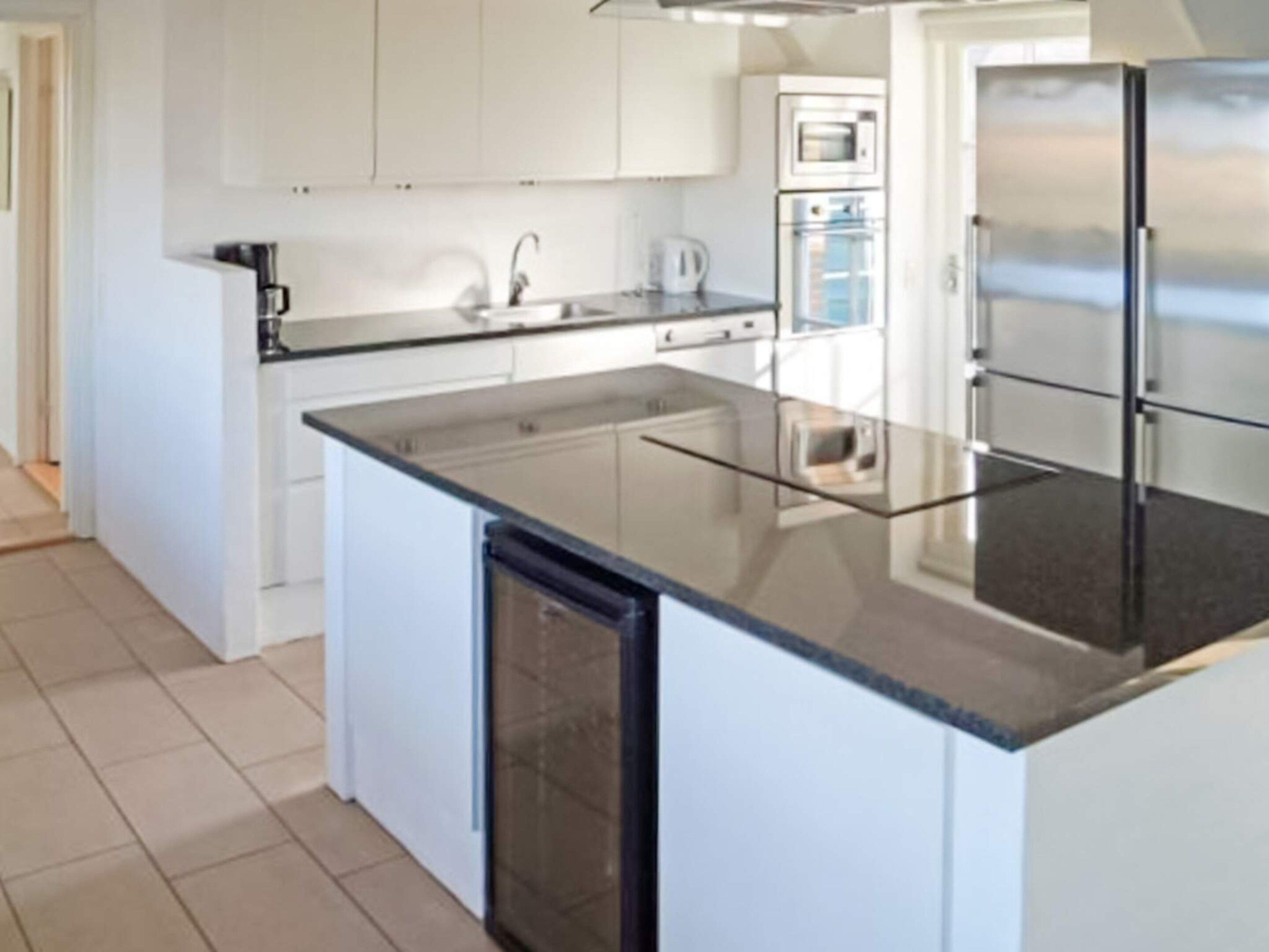 Cozinha moderna com armários brancos, bancada ilha em vidro, eletrodomésticos de aço inoxidável e lava-louças.