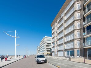 Uma estrada costeira com um carro branco, passarela para pedestres e edifícios altos com varandas.