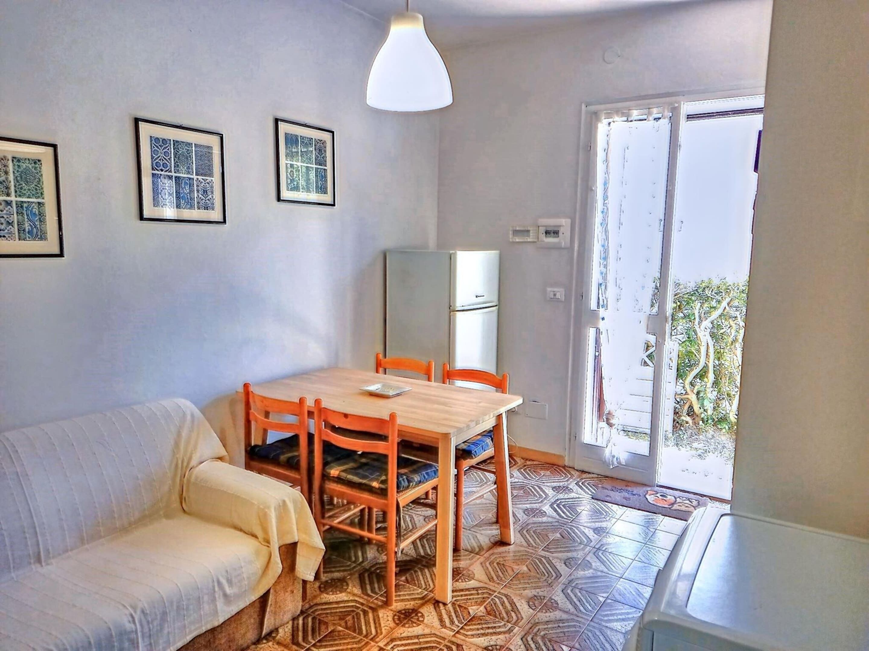 Um quarto pequeno e bem iluminado, com uma mesa de jantar de madeira e cadeiras, um sofá e uma geladeira.