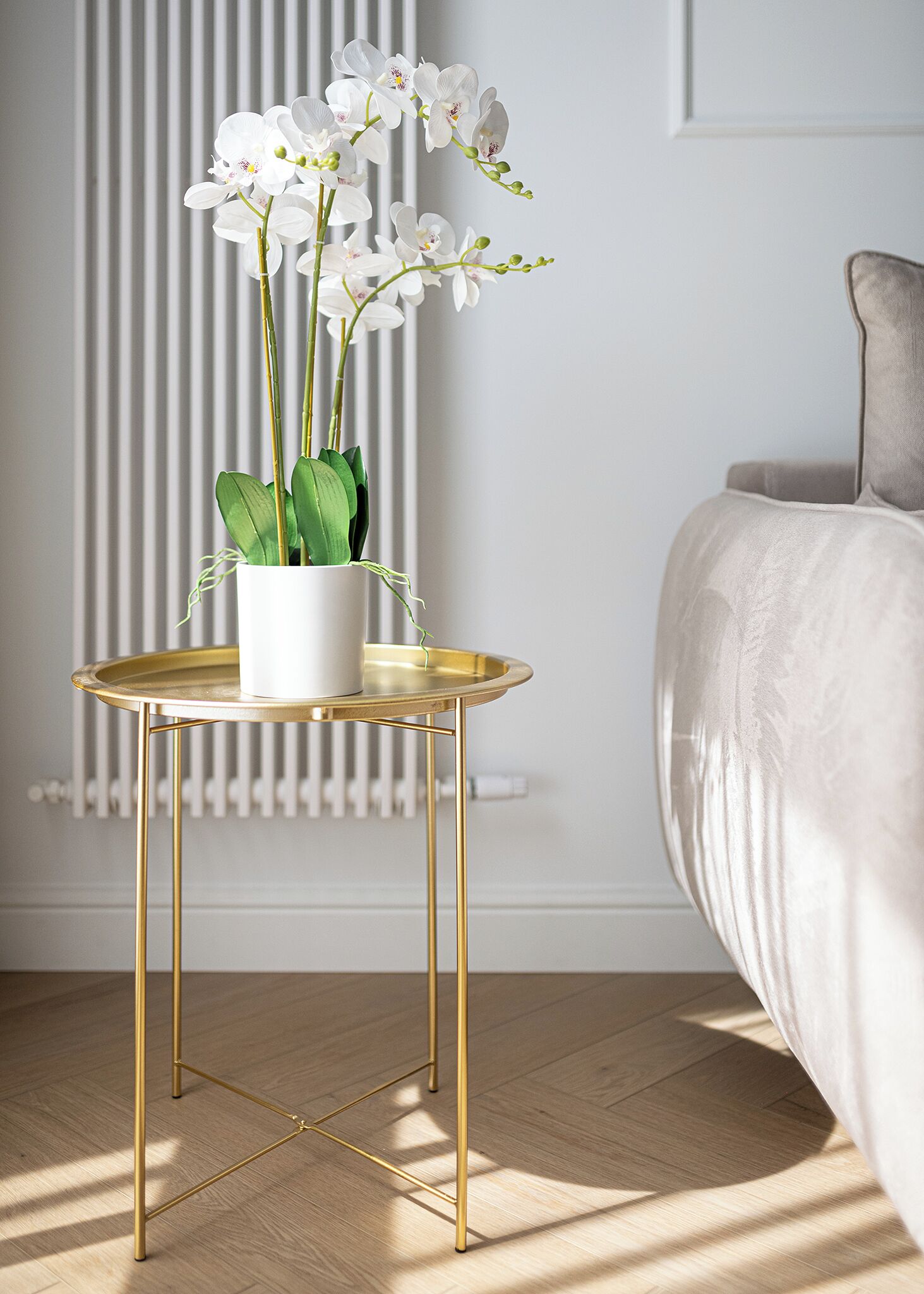 Uma orquídea branca num vaso branco sobre uma mesa dourada e branca, com um radiador ao fundo.