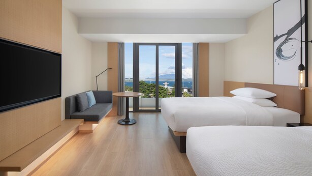 Quarto de hotel moderno com uma janela grande, um sofá, duas camas e uma televisão de tela plana.