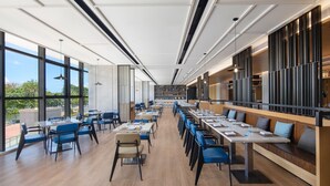 Um restaurante moderno com mesas compridas e cadeiras azuis, janelas amplas e vista para o exterior.