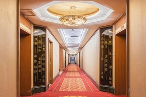 Corredor de hotel com carpete vermelho, portas de madeira e um candelabro.