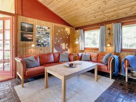 Interior acolhedor de uma cabana, com teto de madeira, sofá de couro, mesa de centro de madeira e uma janela grande com cortinas.