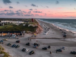 Uma estrada costeira com carros estacionados, uma praia e um edifício com uma estrutura vermelha numa colina.