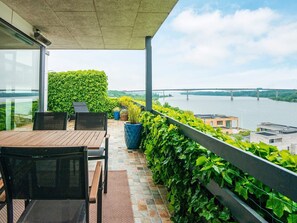 Uma varanda com mesa e cadeiras, uma sebe verde e vista para um rio e uma ponte.