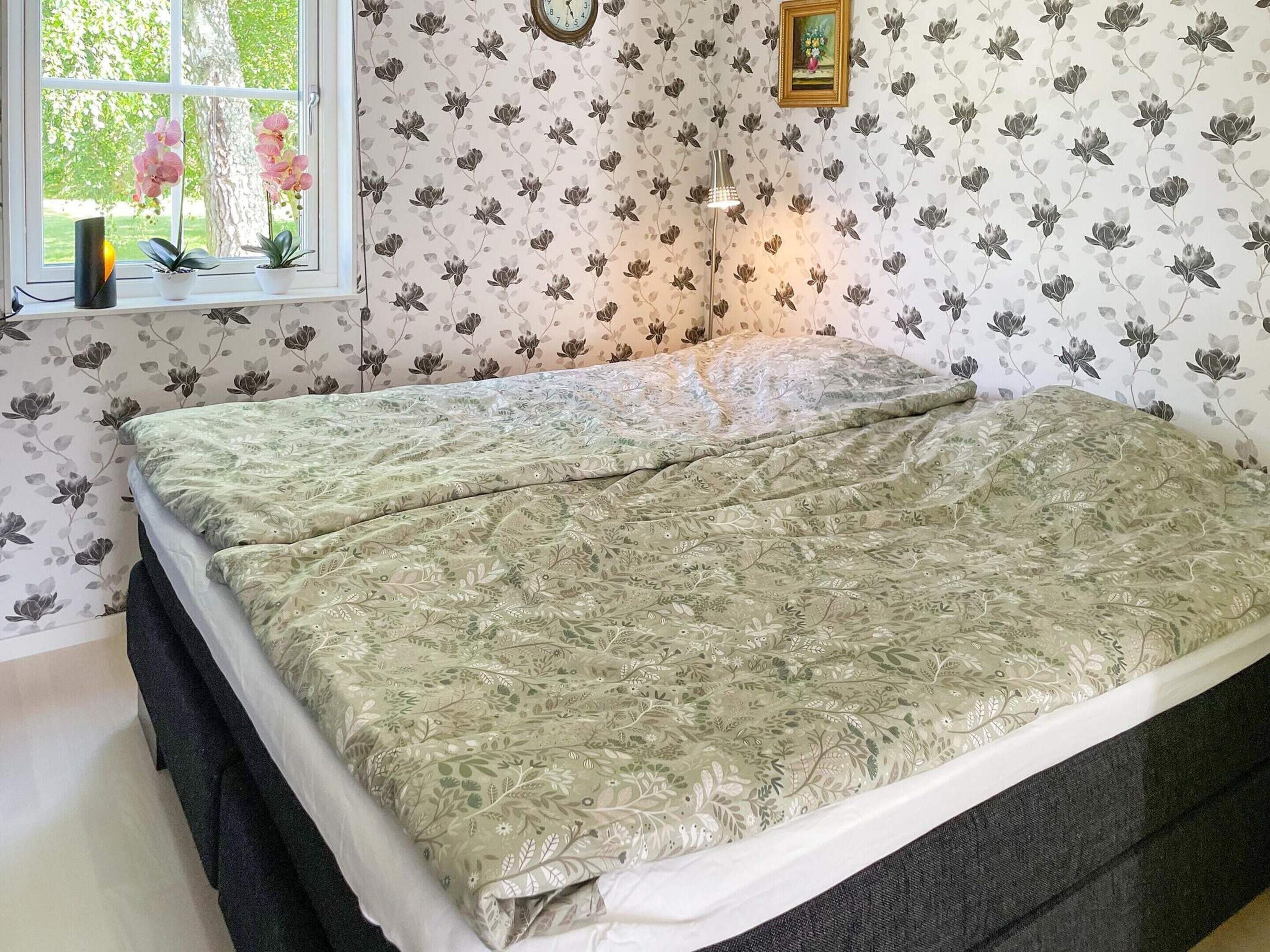 Quarto com cama, papel de parede floral, janela com plantas e relógio de parede.