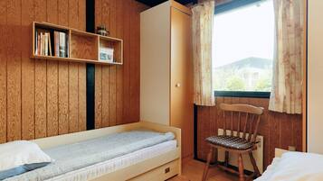 Quarto com uma cama de solteiro, uma estante de madeira, uma cadeira e uma janela com cortinas.