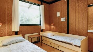 Quarto pequeno e acolhedor com paredes de madeira e uma janela grande. Há duas camas, uma de solteiro e outra de casal. Uma pequena mesa está colocada entre as camas.