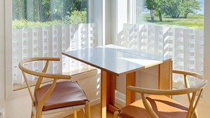 Uma pequena sala de jantar com uma mesa e duas cadeiras, uma janela com vista para vegetação e um separador branco com aberturas quadradas.