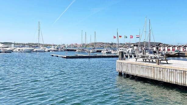 Uma marina com barcos atracados, um cais de madeira e bandeiras ao fundo.
