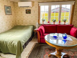 Um quarto com uma cama, um sofá vermelho, uma mesa de vidro redonda e uma janela com plantas em vasos.