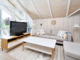 Uma sala de estar moderna com uma TV sobre um suporte de madeira, um sofá cinzento, uma mesa de centro branca e uma grande janela que proporciona luz natural.