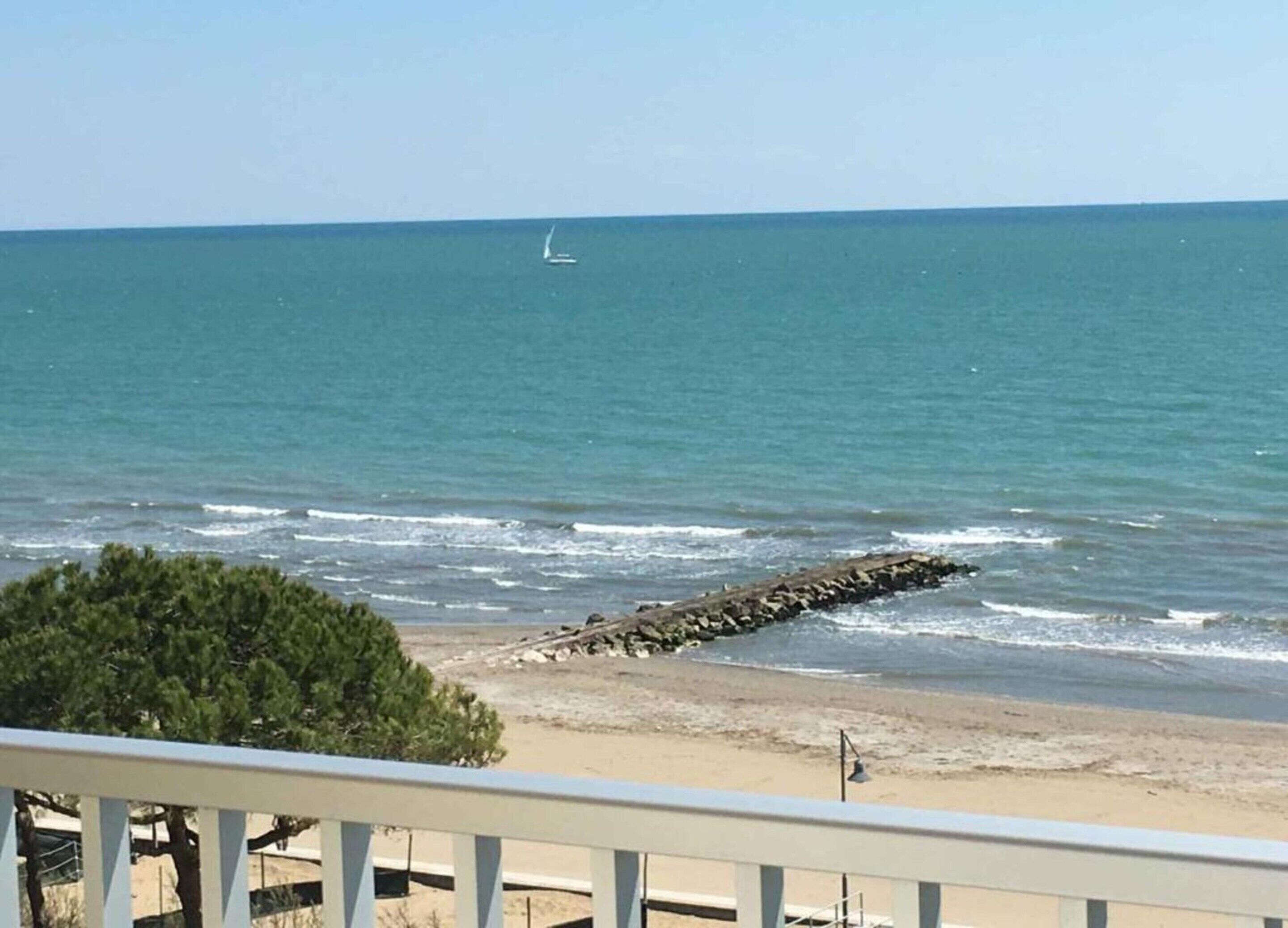 Uma praia com um molhe rochoso, um veleiro ao longe e um céu claro.