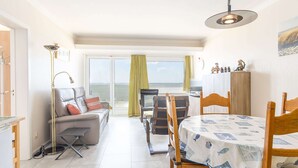 Quarto com mesa de jantar, cadeiras, sofá, televisão e vista para o mar através de uma grande janela.