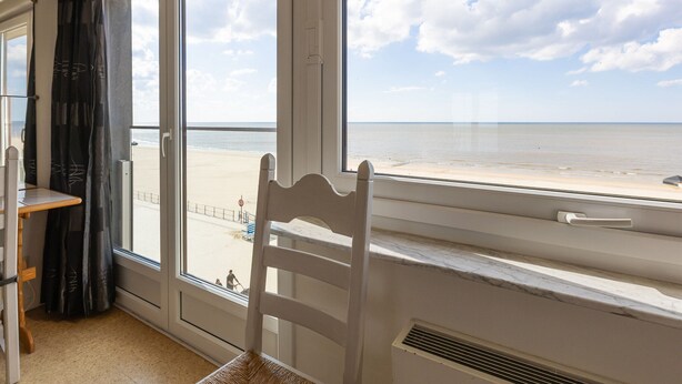 Quarto com uma janela grande de frente para uma praia, uma cadeira e uma pequena mesa.