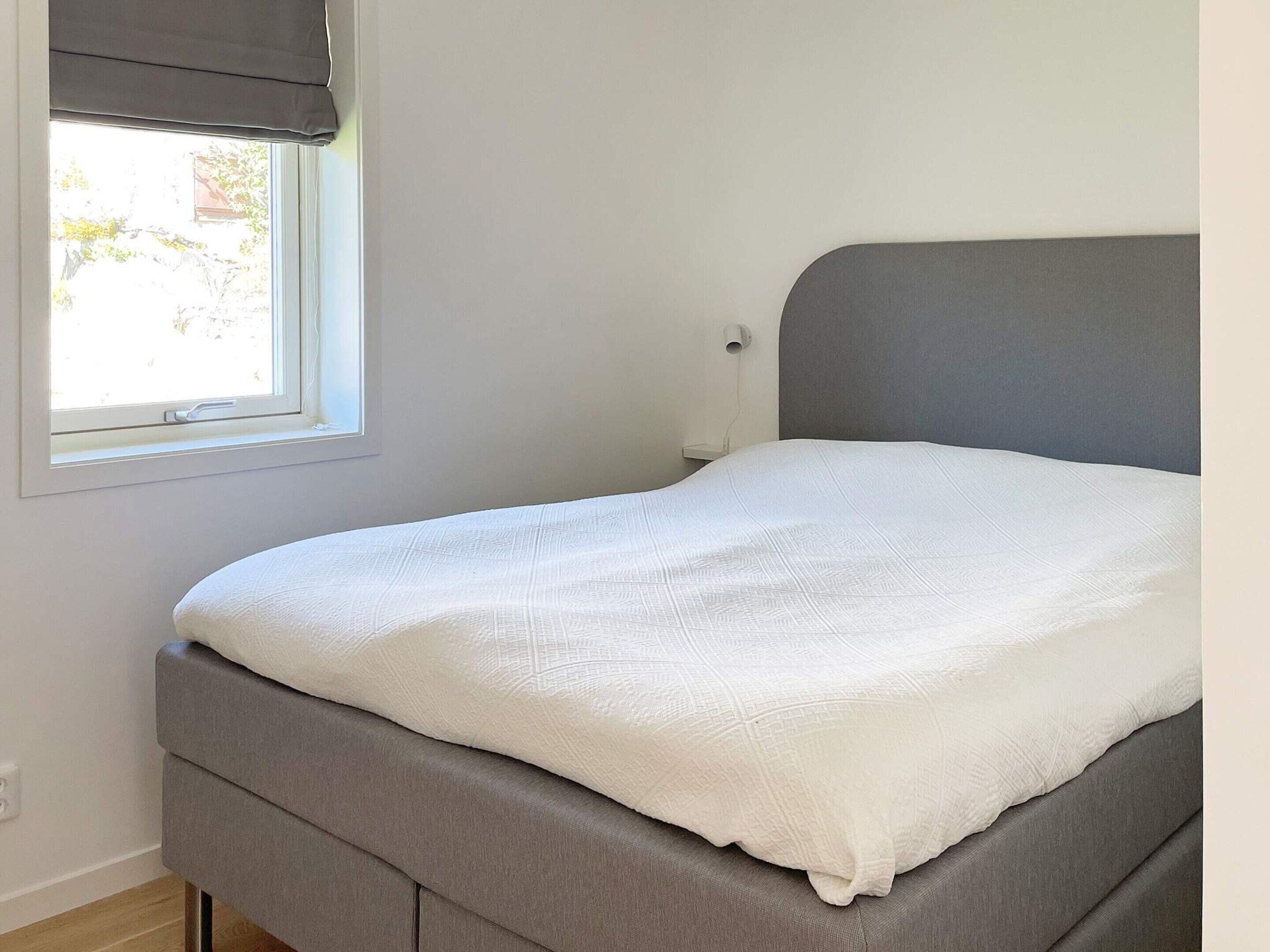 Cama com cabeceira cinzenta e manta branca num quarto com janela e tomada.