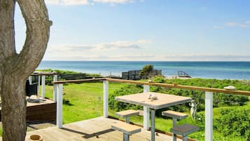 Um deck de madeira com mesa e bancos, com vista para uma área gramada e o mar.