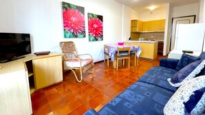 Uma sala de estar com sofá, televisão, mesa de jantar com cadeiras e área de cozinha.