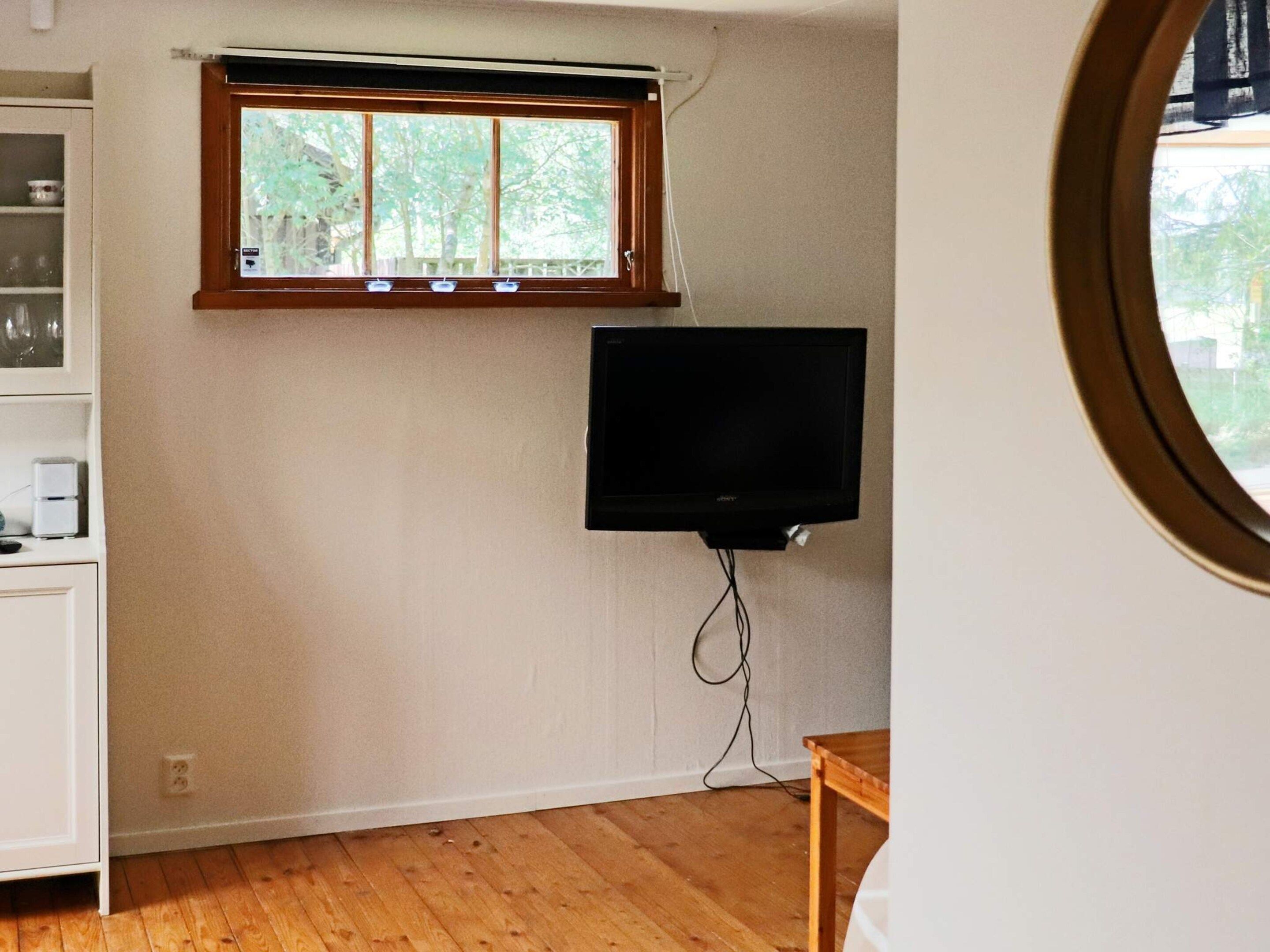 Quarto com piso de madeira, janela com vista para árvores, TV de tela plana na parede e uma pequena mesa de madeira.