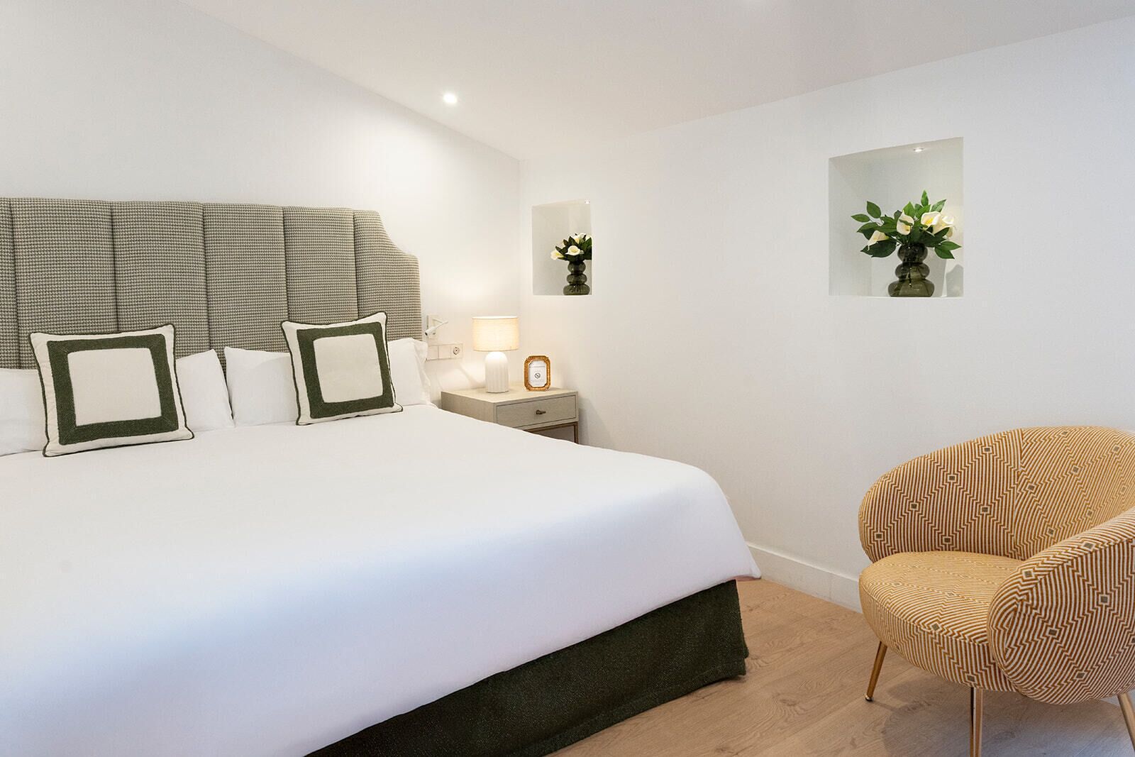 Quarto de hotel com uma cama grande, uma mesa de cabeceira com abajur, uma cadeira e uma pequena planta na parede.