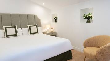 Quarto de hotel com uma cama grande, uma mesa de cabeceira com abajur, uma cadeira e uma pequena planta na parede.
